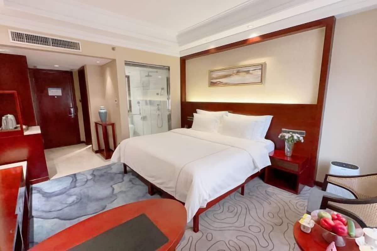 Quarto de hotel com uma cama grande, uma escrivaninha, uma cadeira, uma mesinha e um banheiro visível por uma porta de vidro.