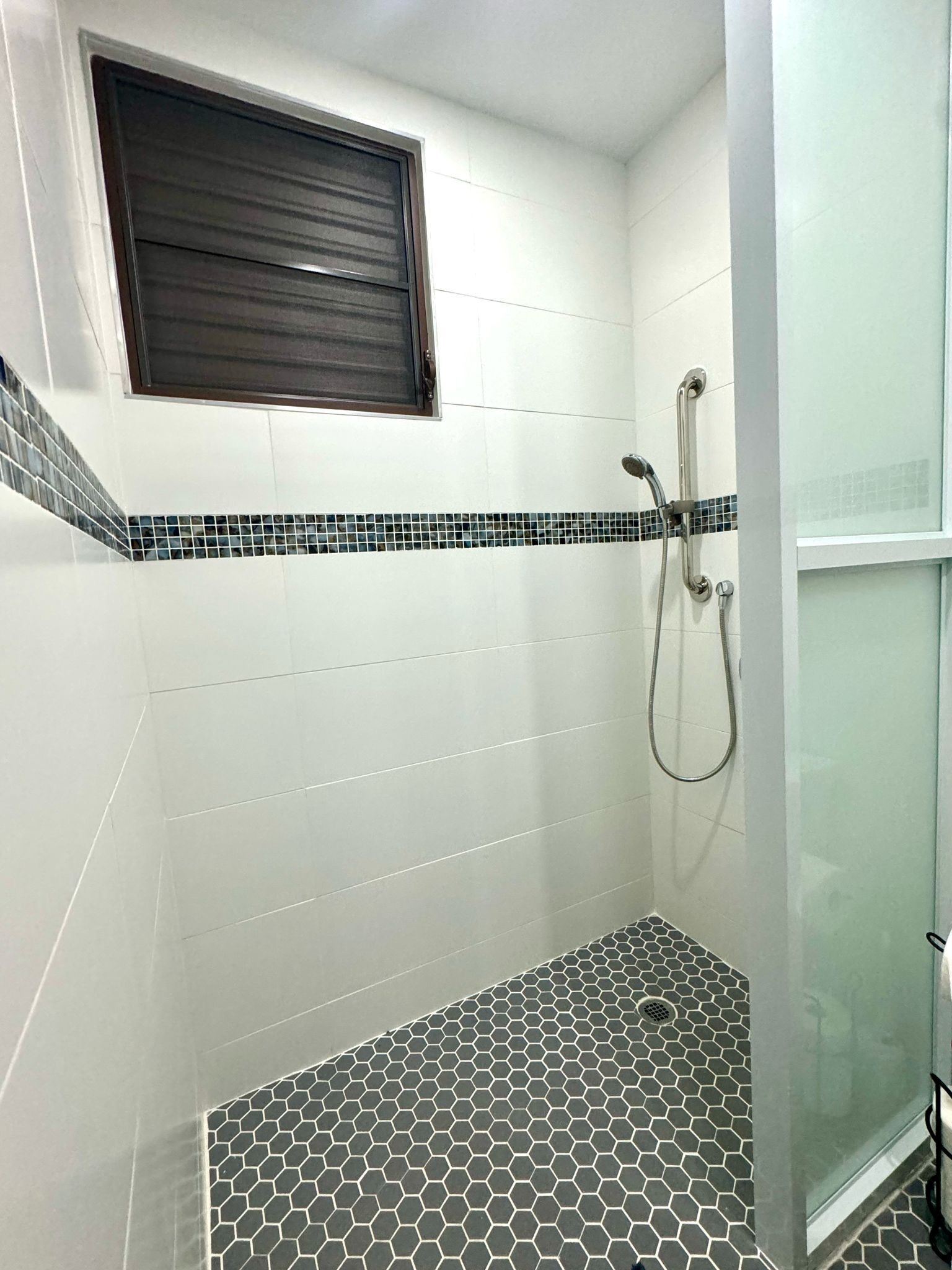 Área de chuveiro com paredes revestidas de azulejos brancos, uma pequena janela e piso em azulejos hexagonais.