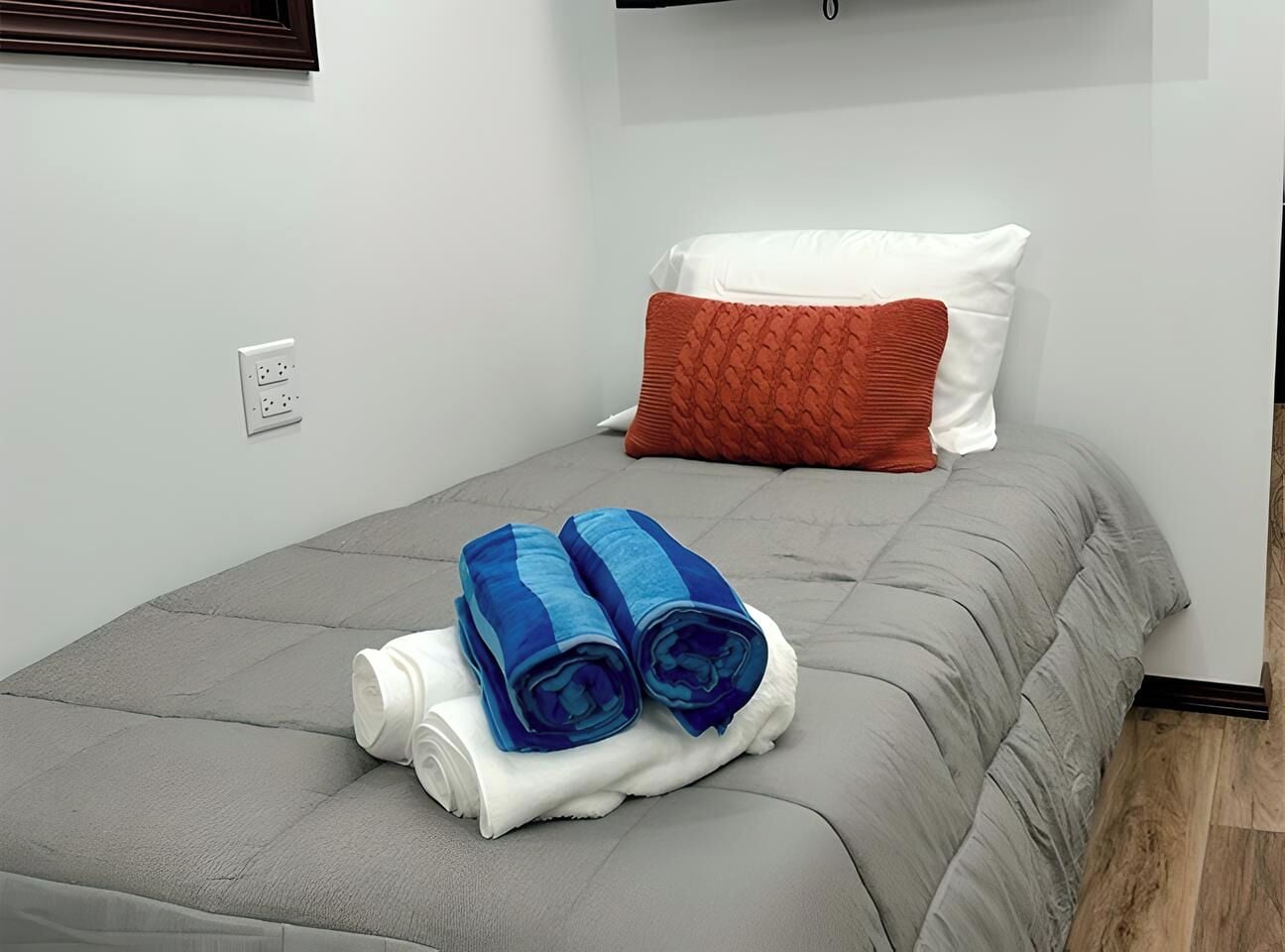 Cama bem arrumada com roupa de cama cinza, um travesseiro branco e um travesseiro vermelho. Há duas toalhas enroladas na cama, uma branca e outra azul.