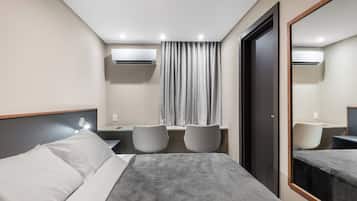 Quarto de hotel com cama, duas cadeiras, escrivaninha, espelho e ar condicionado.