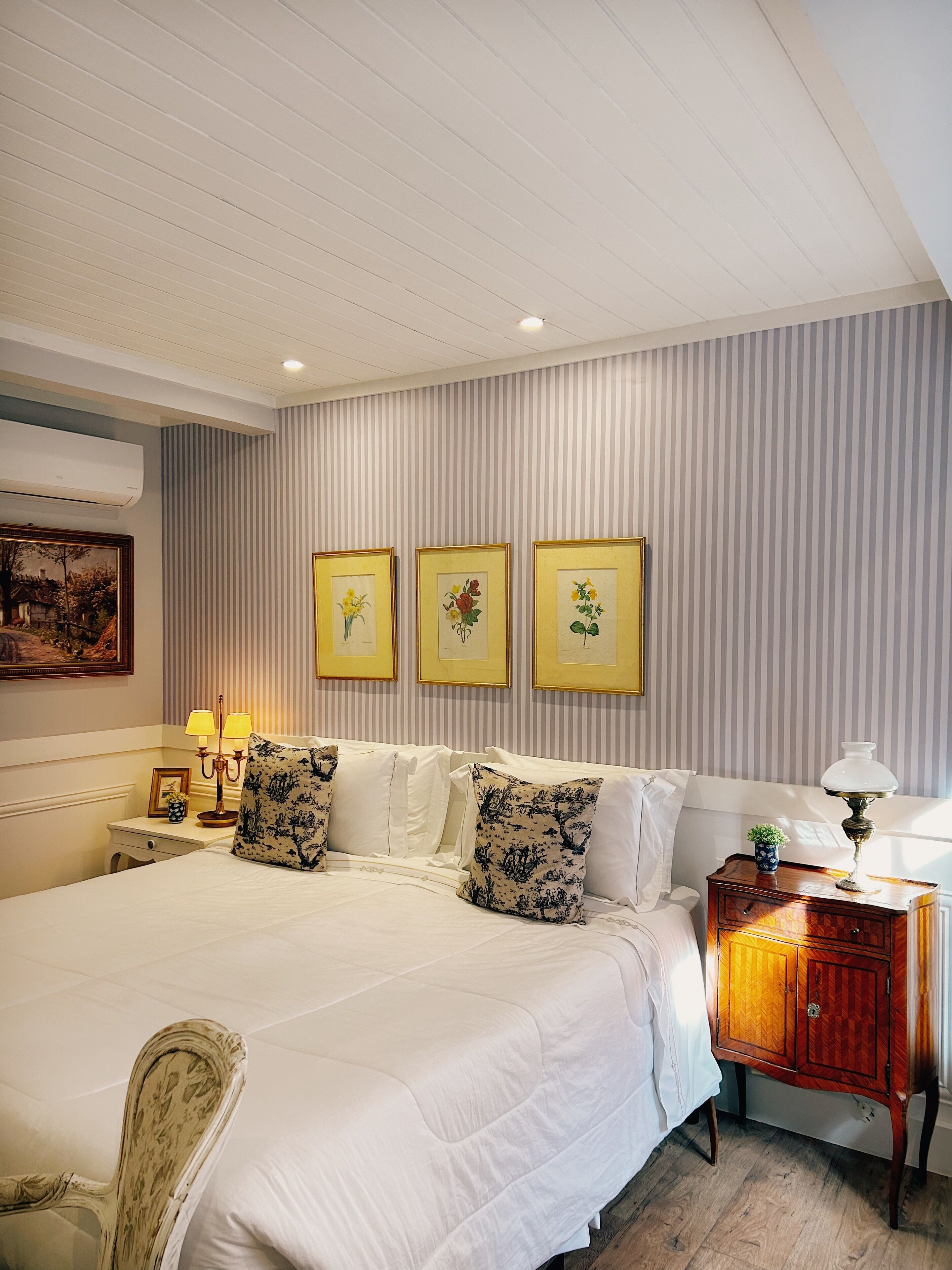 Quarto de hotel com uma cama grande, uma mesa de cabeceira com abajur e três quadros na parede.