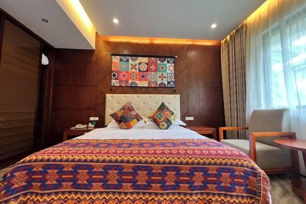 Quarto de hotel com uma cama grande, cabeceira com um colcha colorida, mesa de cabeceira pequena, uma cadeira e uma janela com cortinas.