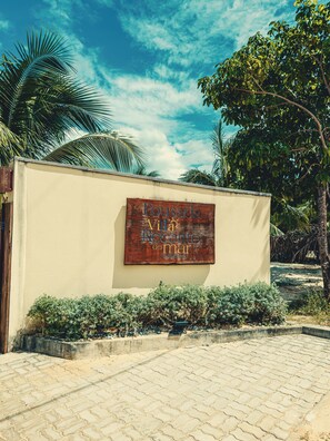 Placa indicando 'Villa Recanto do mar', com entrada pavimentada e vegetação tropical.