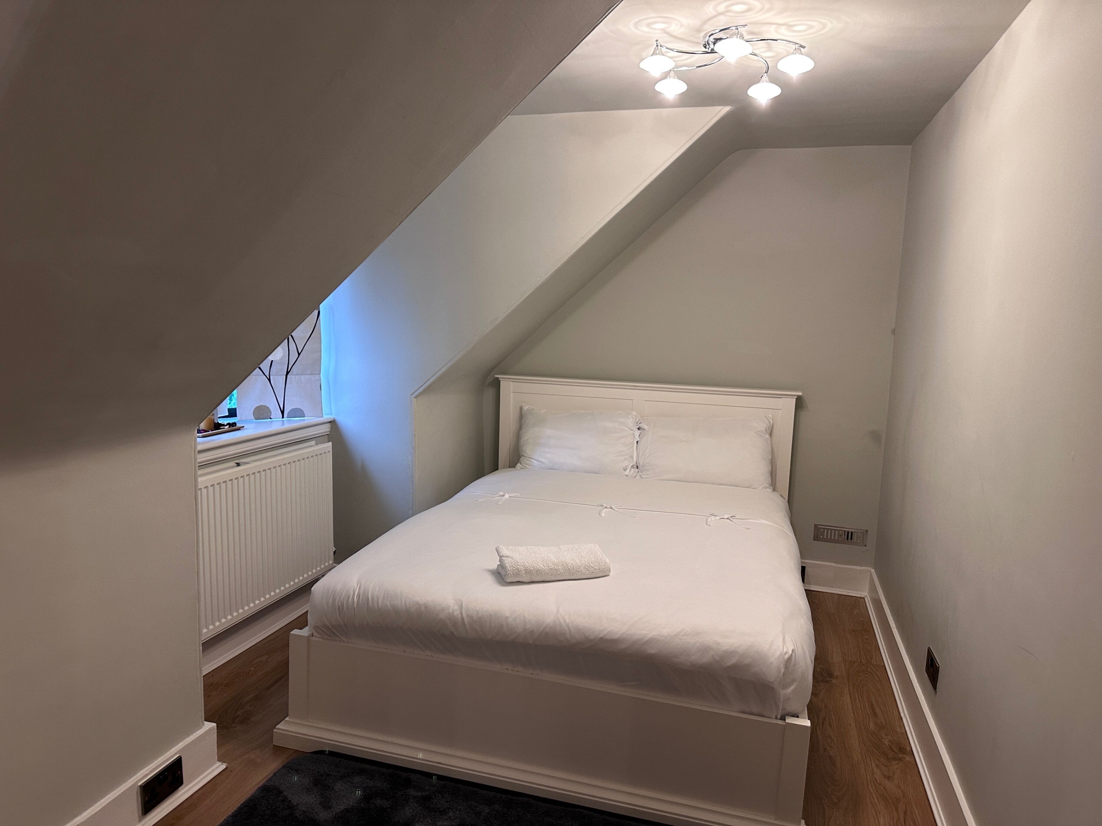 Quarto com cama branca, radiador, tapete escuro e teto inclinado com luminária.