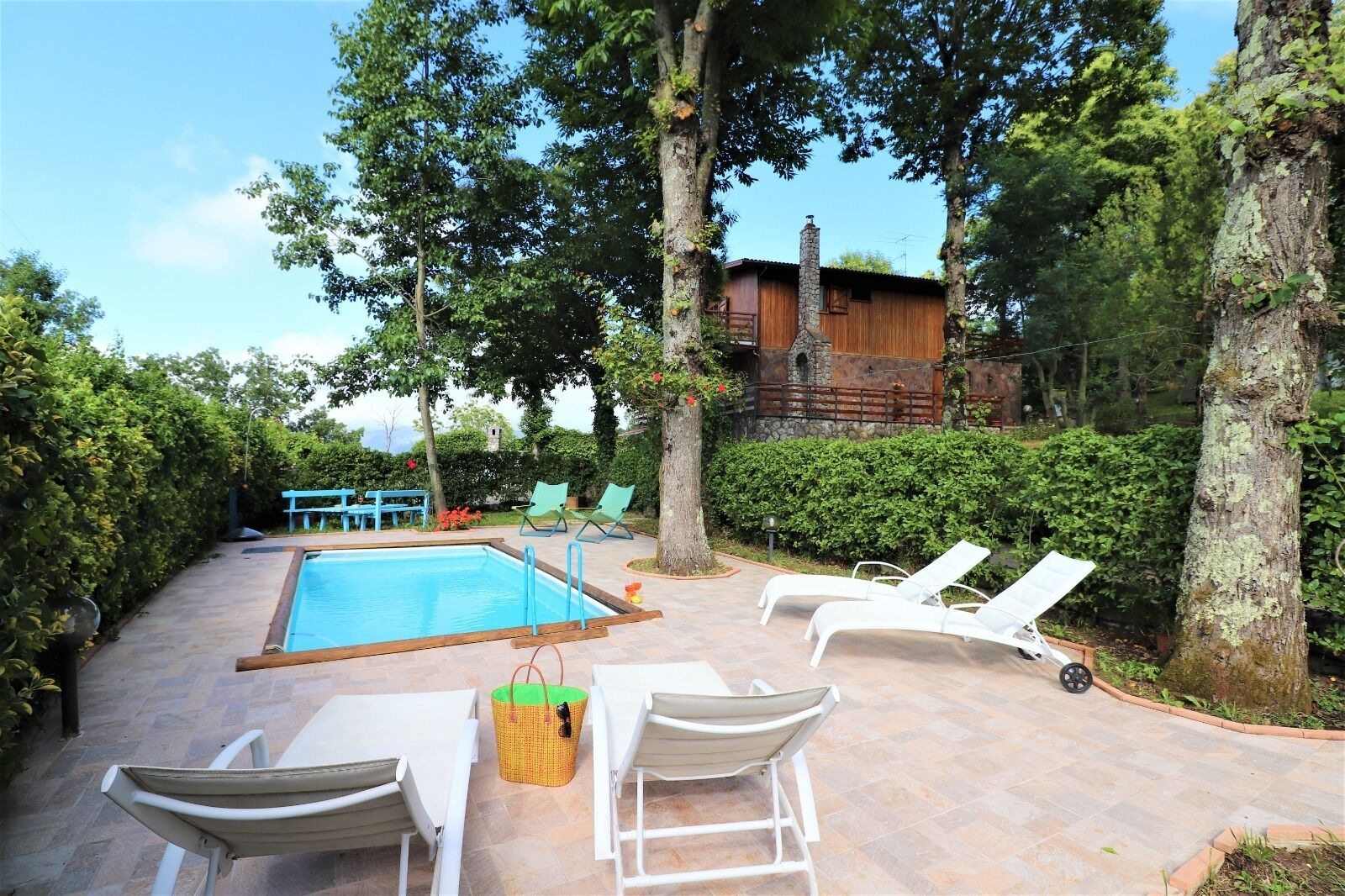Piscina com espreguiçadeiras e uma mesa, cercada por árvores e um prédio de madeira.