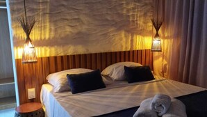 Quarto de hotel com uma cama grande, duas luminárias fixadas na parede, um banquinho redondo e uma toalha enrolada sobre a cama.