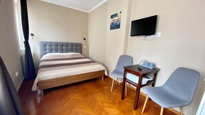 Quarto de hotel com cama, televisão, escrivaninha com cadeiras e piso de madeira.