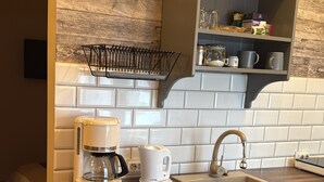 Uma cozinha compacta com painel de madeira, armários brancos, máquina de café, chaleira e pia.