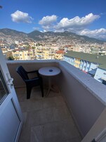 Uma varanda com uma mesa redonda e uma cadeira, com vista para uma paisagem urbana com edifícios e montanhas.