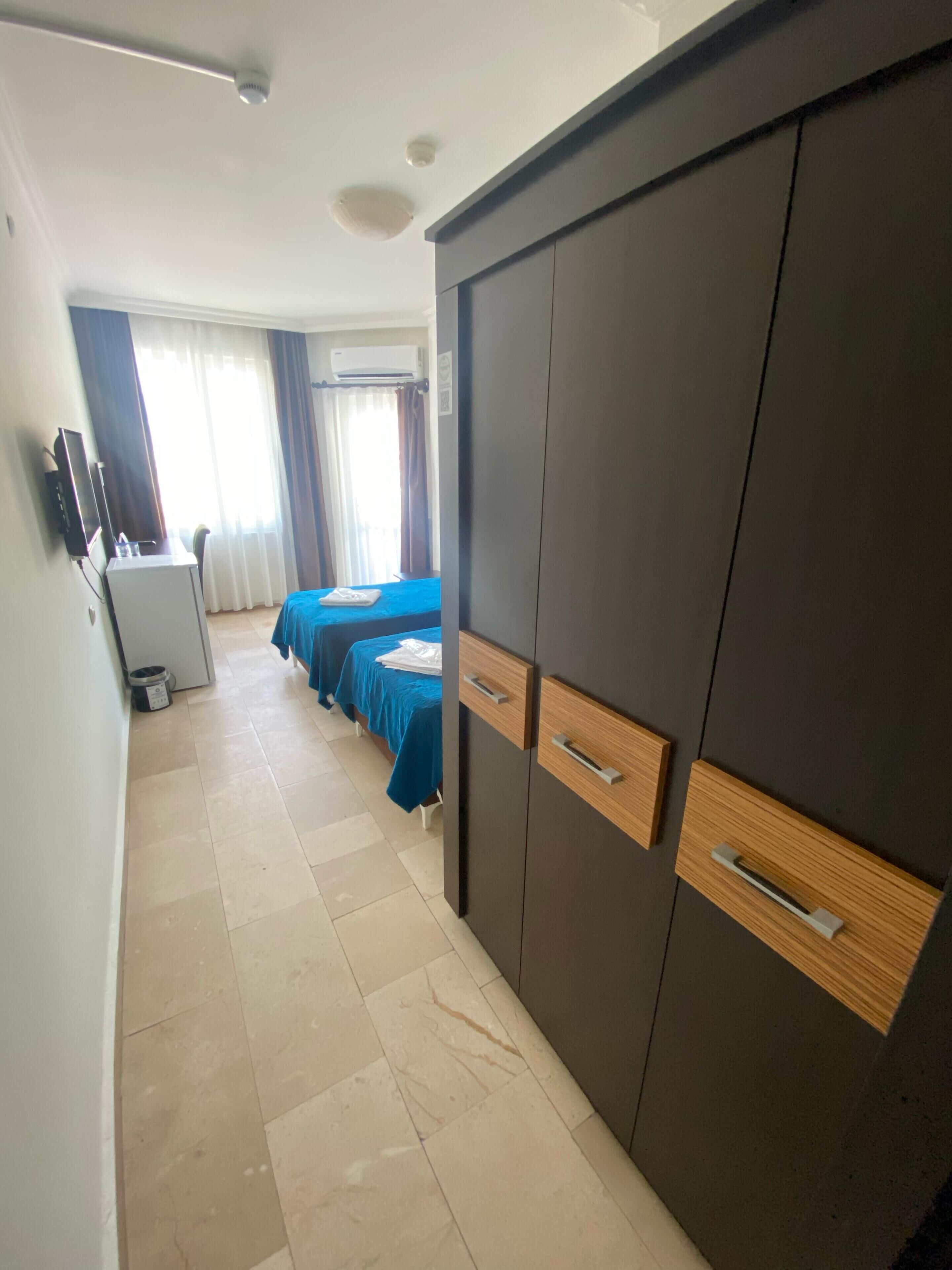 Quarto de hotel com um guarda-roupa grande, uma escrivaninha com cadeira, duas camas com cobertores azuis e um aparelho de ar condicionado.