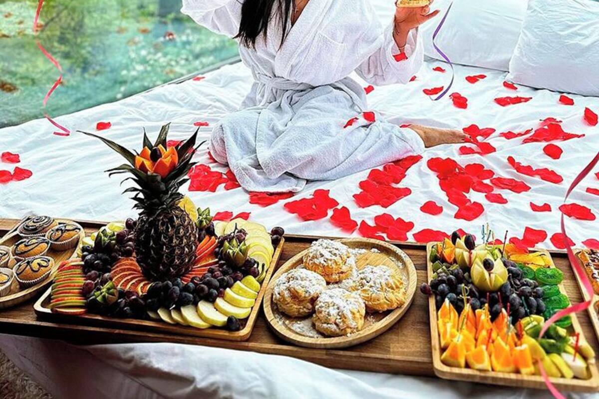 Uma pessoa vestida com um robe branco sentada em uma cama, com vista panorâmica, cercada por frutas e doces.