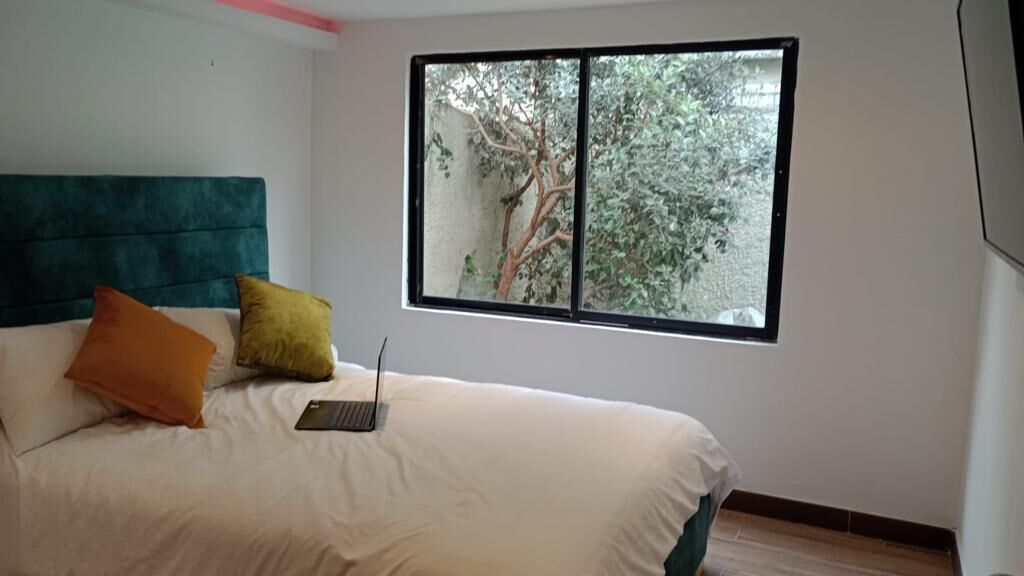 Quarto com cama, almofadas, um laptop e janela com vista para árvores.