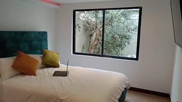 Quarto com cama, almofadas, um laptop e janela com vista para árvores.