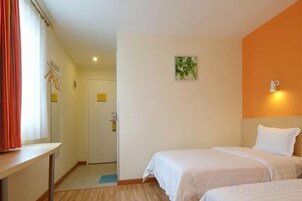 Quarto de hotel com duas camas, uma escrivaninha, uma porta e uma pequena janela com cortinas.