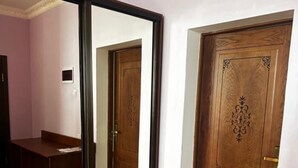 Quarto com piso de madeira, um espelho grande, porta de madeira com detalhes entalhados e uma pequena mesa com uma toalha.