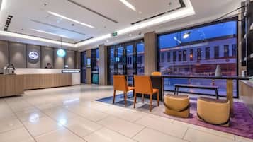 Uma área de recepção de hotel, com balcão, espaço de estar e grandes janelas com vista para a rua.