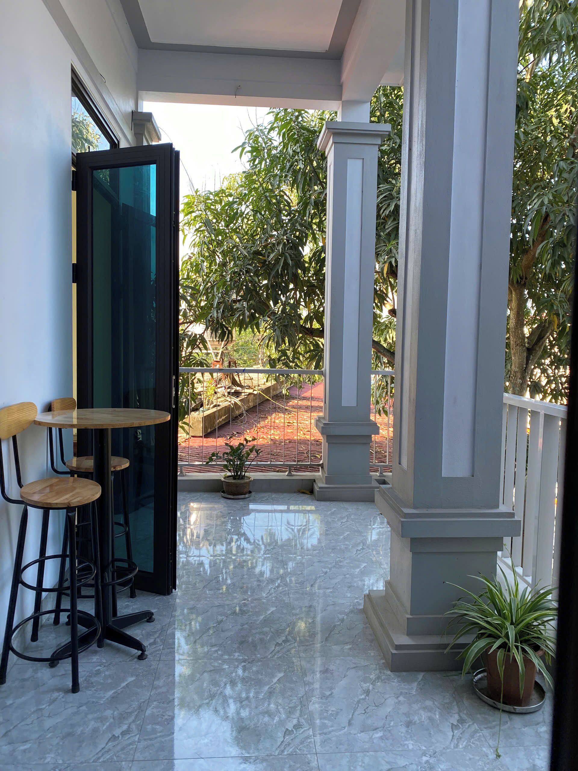 Uma varanda com porta de vidro, uma mesa redonda pequena, duas cadeiras de madeira e um vaso com planta.