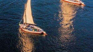 Cena costeira com veleiros em águas calmas, uma marina com barcos atracados e ao fundo uma paisagem acidentada com edifícios e árvores.