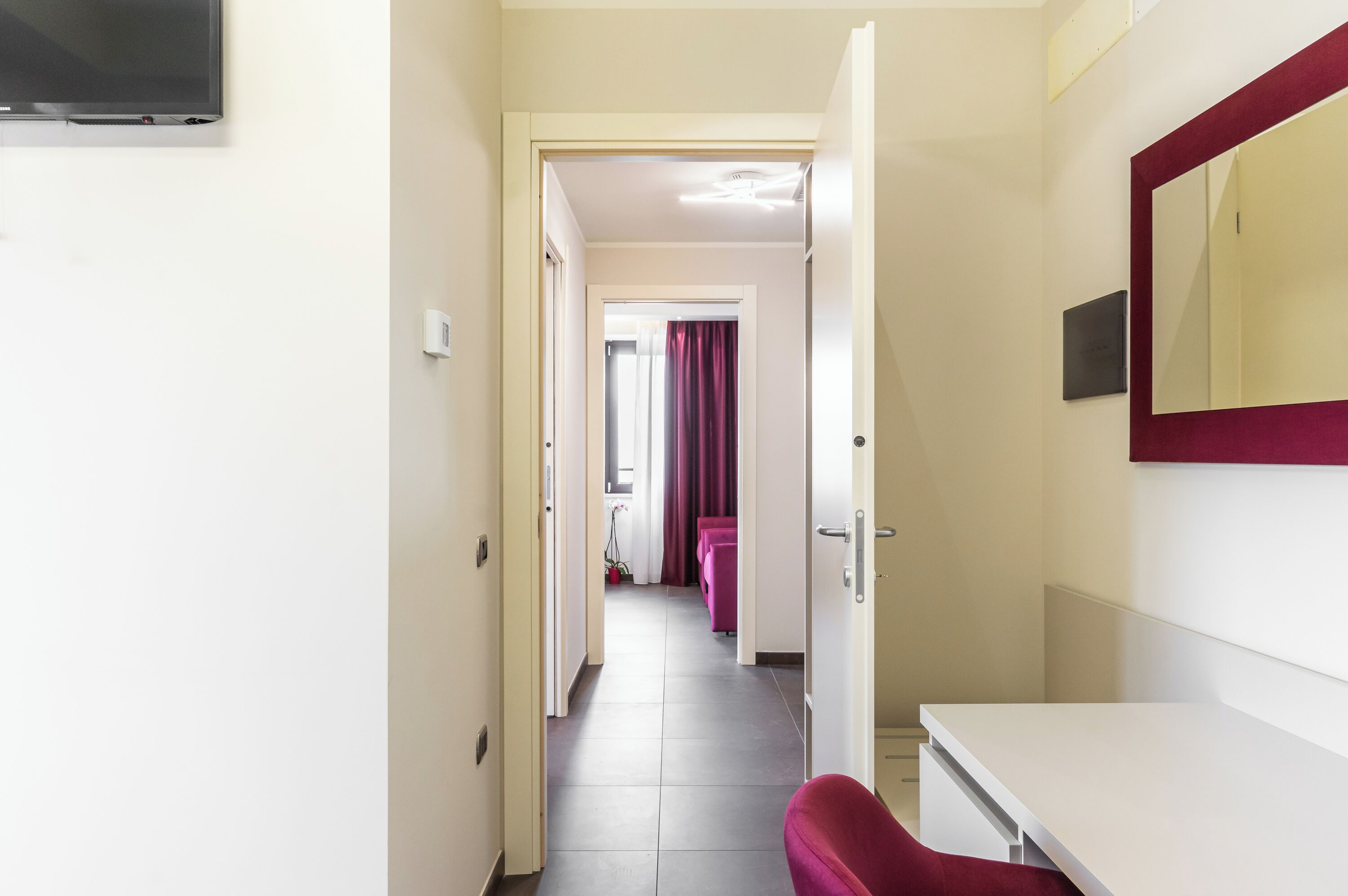 Um corredor de hotel com parede branca, uma televisão de tela plana montada acima de uma porta, um espelho com moldura vermelha e uma cadeira vermelha no quarto.