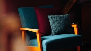 Uma cadeira de madeira com almofada azul e travesseiro vermelho num quarto com iluminação suave e soalho de madeira.