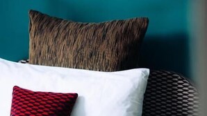 Cama com roupa de cama branca e duas almofadas, uma vermelha e outra marrom, em frente a uma parede verde-água.
