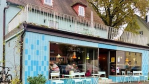 Um café com mesas ao ar livre, uma bicicleta estacionada na frente e um vaso de planta em primeiro plano.