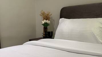 Uma cama bem arrumada com lençóis brancos, cabeceira escura e um pequeno vaso com flores secas em uma mesa de cabeceira.