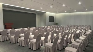 Sala de conferências com fileiras de cadeiras e mesas, um pódio e uma parede em tom neutro.