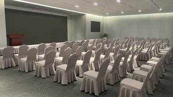 Sala de conferências com fileiras de cadeiras e mesas, um púlpito e esquema de cores neutras.