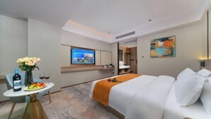 Quarto de hotel com cama, televisão, pequena mesa com garrafa e flores, e um quadro na parede.