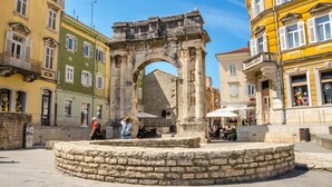 Uma antiga arcada de pedra numa praça pública, com edifícios circundantes e áreas de estar ao ar livre.