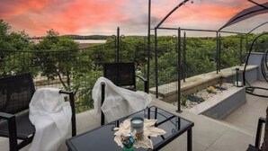 Terraço ao ar livre com mesa e cadeiras, vista para vegetação e água, e céu ao pôr do sol.