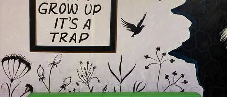 Um sofá verde num quarto com um mural de aves e plantas, e uma placa com a inscrição 'DON'T GROW UP IT'S A TRAP'.