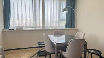 Quarto de hotel com uma mesa de jantar e cadeiras, uma pequena mesa lateral, um abajur e vista da cidade através de cortinas finas.