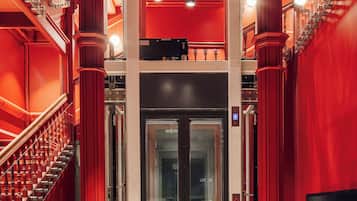 Entrada de elevador com paredes vermelhas, porta de vidro, escada à esquerda e um pilar vermelho à direita.