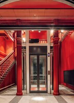Entrada de elevador com paredes vermelhas, porta de vidro, escada à esquerda e um pilar vermelho à direita.