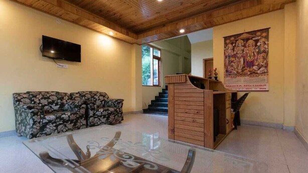 Uma sala de estar com um sofá floral, uma mesa de centro de vidro, uma escada de madeira e uma televisão fixada na parede.