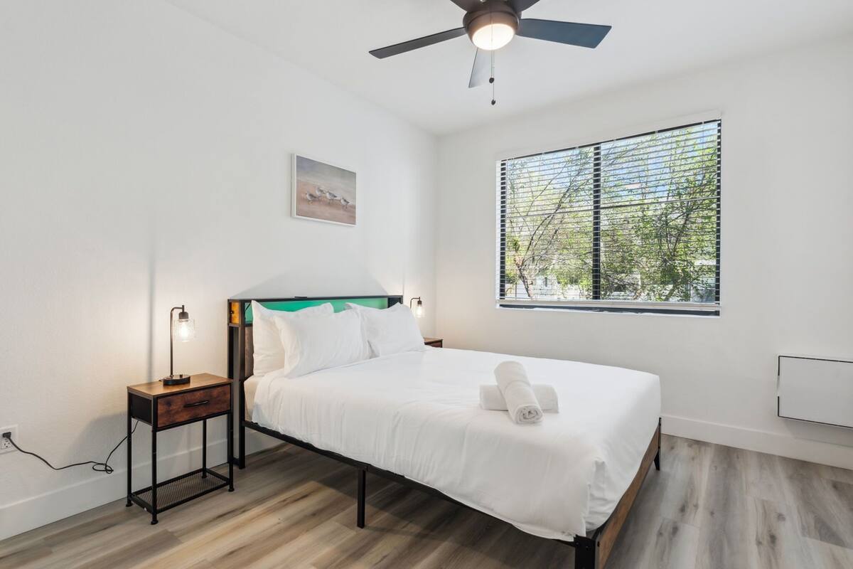Quarto moderno com cama, mesa de cabeceira, ventilador de teto, janela com persianas e um quadro na parede.