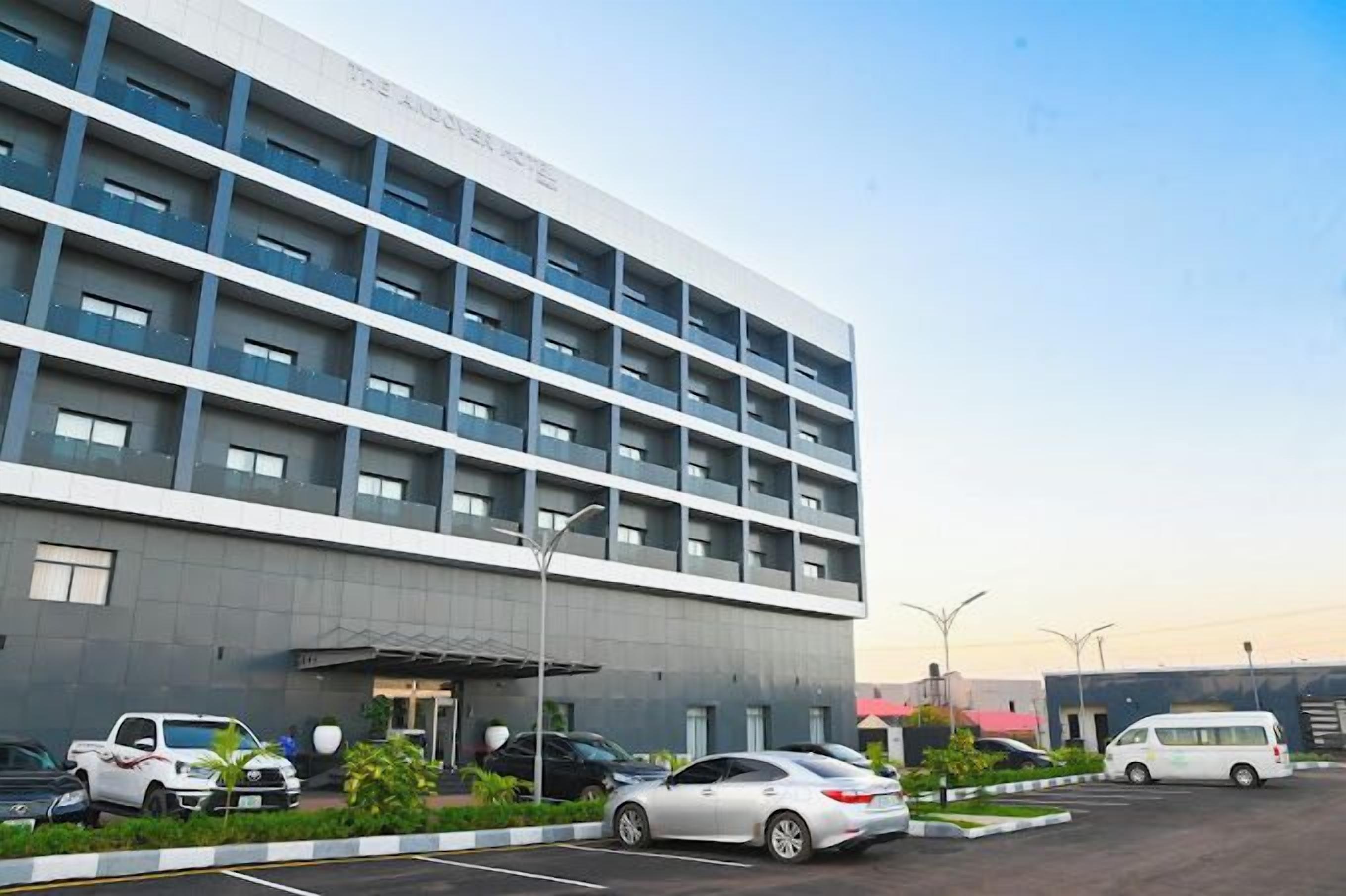 Hotel de vários andares com design moderno, apresentando um estacionamento com diversos carros e uma pequena área ajardinada.