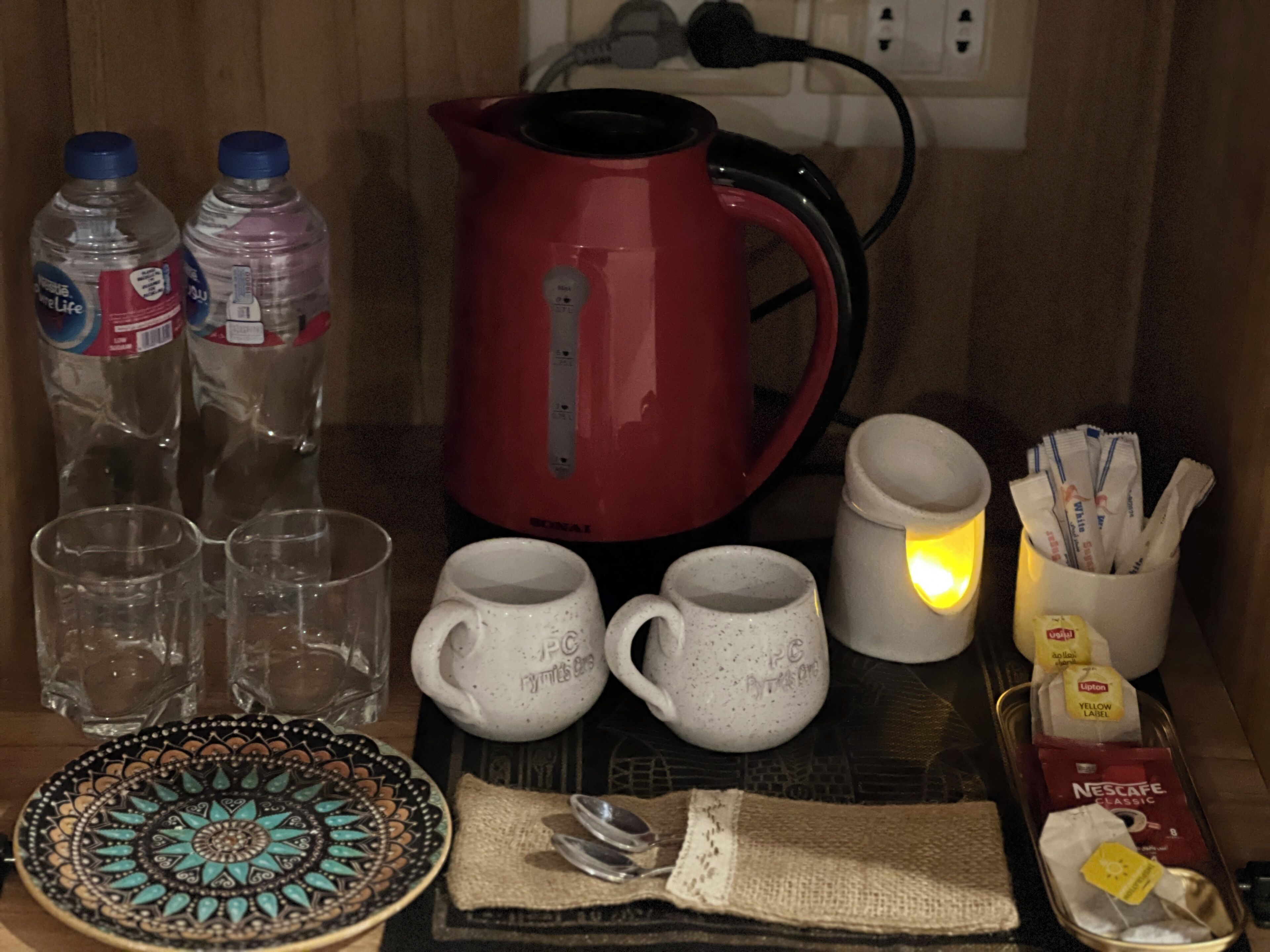 Uma bancada de cozinha com uma chaleira vermelha, duas garrafas de água, dois copos, duas canecas brancas, uma vela acesa, um prato decorativo, um guardanapo, uma colher, uma embalagem de chá Lipton e uma embalagem de café Nescafé Classic.
