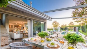 Uma área de refeições moderna ao ar livre, com piscina, móveis de vime e vista para o jardim.