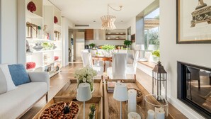 Uma sala de estar moderna com sofá, mesa de centro e lareira. Há uma área de jantar com mesa e cadeiras, e uma cozinha ao fundo.
