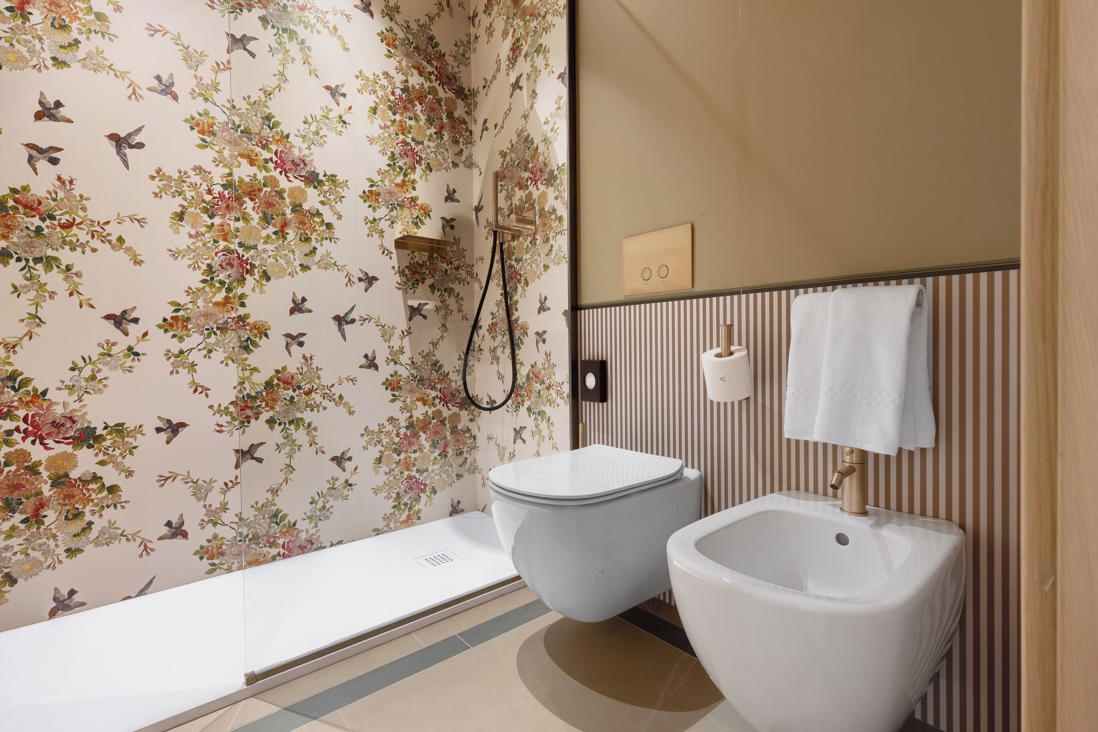 Casa de banho com chuveiro, sanita e lavatório, com papel de parede floral e painéis listrados.