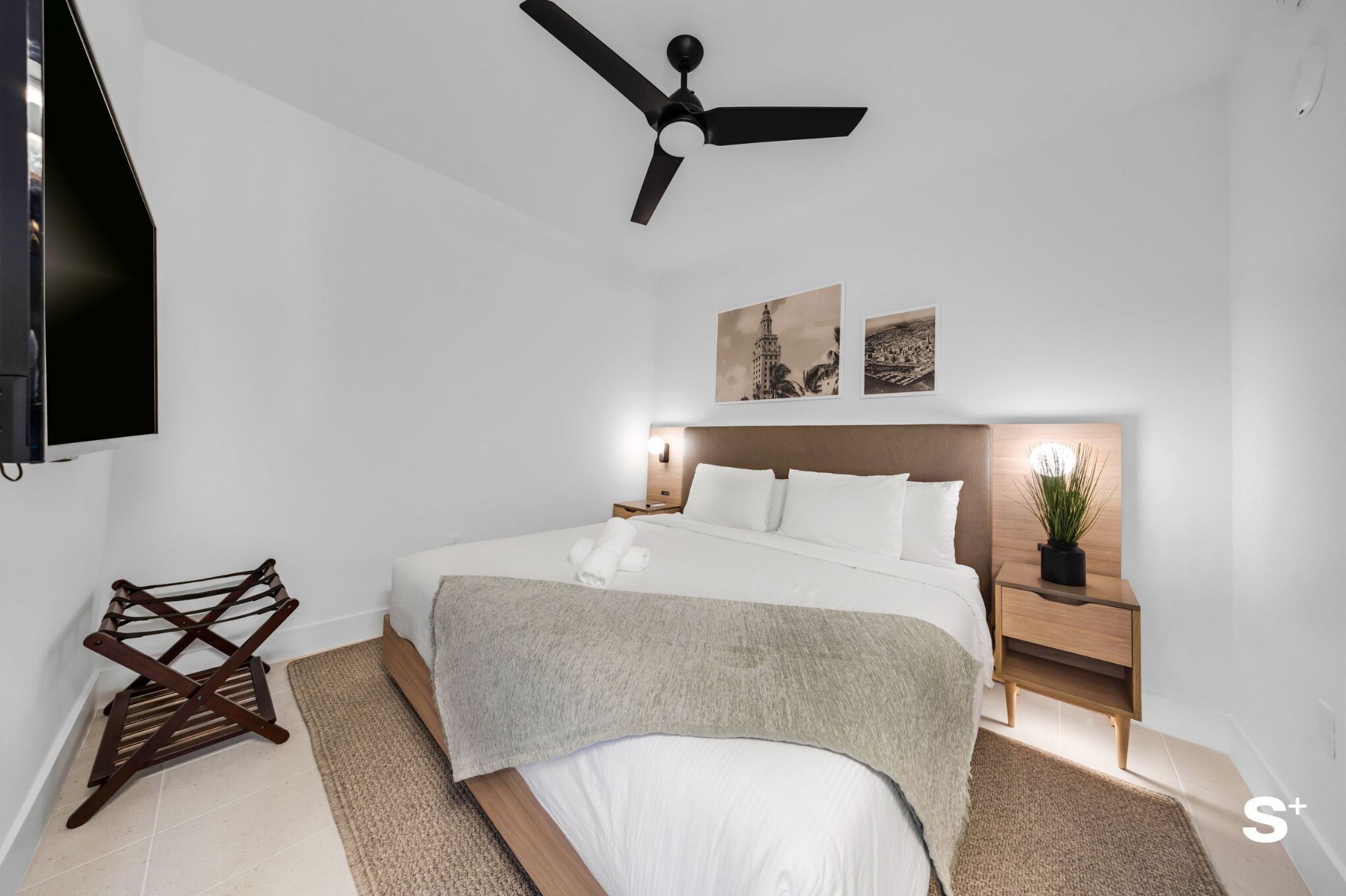 Quarto de hotel moderno com uma cama grande, um ventilador de teto, uma mesa de cabeceira com abajur e duas imagens emolduradas na parede.