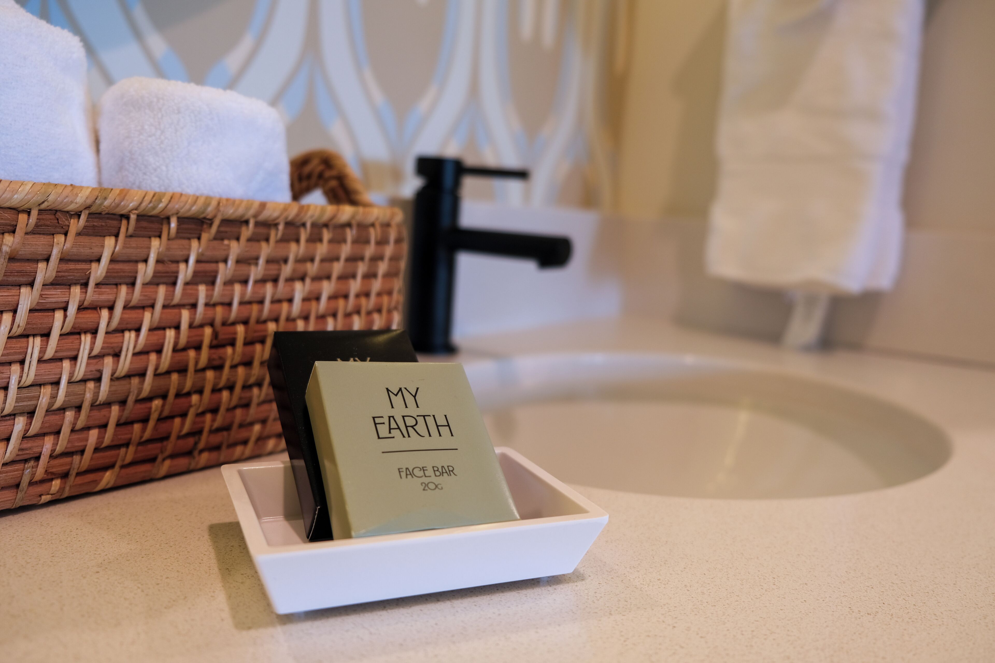 Casa de banho com lavatório branco, cesto de vime com toalhas e um saboneteiro com a etiqueta "MY EARTH FACE BAR 20c".