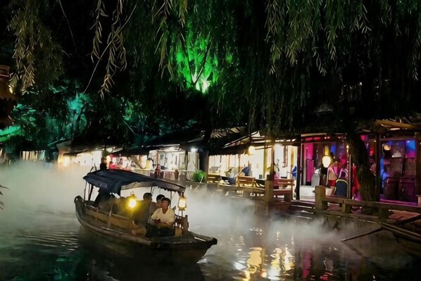 Um barco com passageiros em um rio calmo à noite, cercado por edifícios e árvores iluminados.
