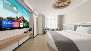 Quarto de hotel moderno com uma televisão de tela plana grande exibindo uma paisagem de montanhas, uma cama com colcha cinza e branca, uma mesa de cabeceira de madeira e uma obra de arte no teto.