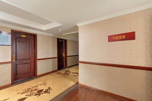 Corredor de hotel com portas de madeira, piso coberto de carpete e placa indicando a direção para a recepção.