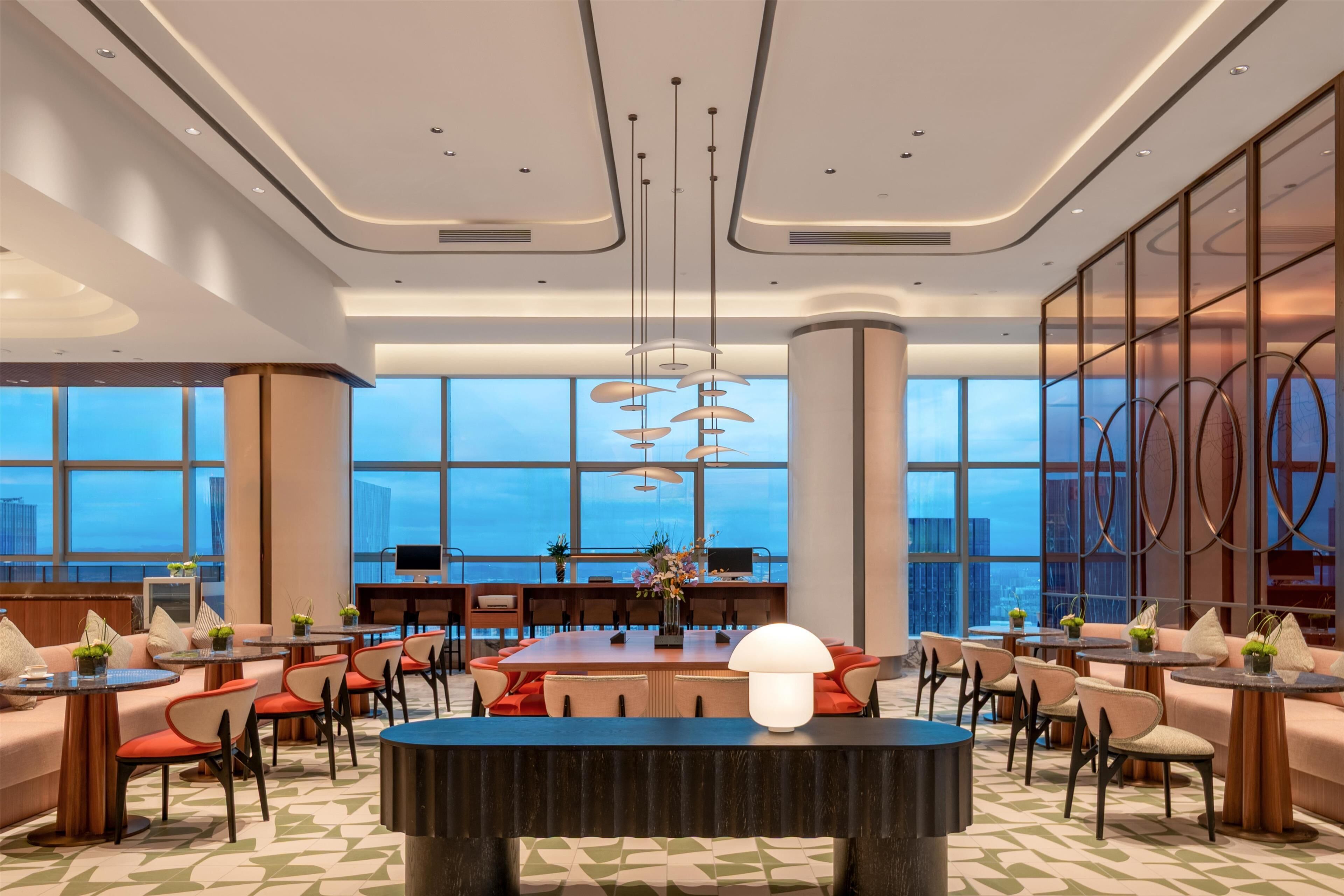 Um saguão de hotel moderno com uma grande mesa de jantar, assentos confortáveis e amplas janelas com vista para a cidade.