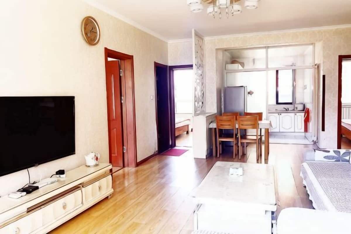 Uma sala de estar com televisão de tela plana, piso de madeira, área de jantar com mesa e cadeiras e cozinha ao fundo.